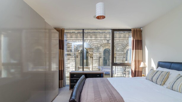Quarto de hotel moderno com uma cama grande, uma escrivaninha e vista para arquitetura histórica através de janelas do chão ao teto.