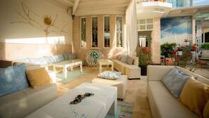 Uma sala de estar ampla, com vários sofás e uma mesa de centro, janelas grandes com cortinas e uma peça decorativa na parede.