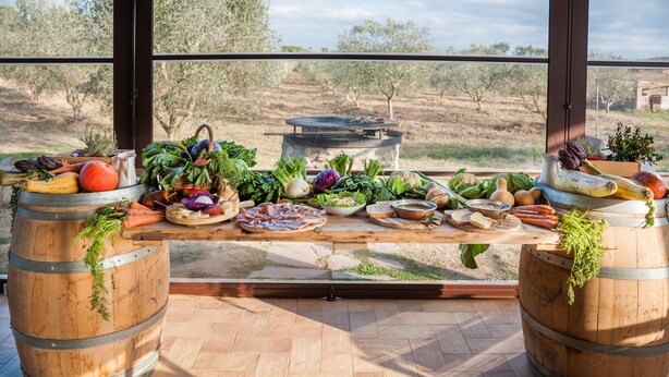 Mesa posta com diversas frutas, legumes e carnes, com vista para um oliveiral através de grandes janelas.