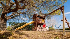 Uma casinha de madeira com escorrega e baloiços, rodeada por árvores e um céu limpo.