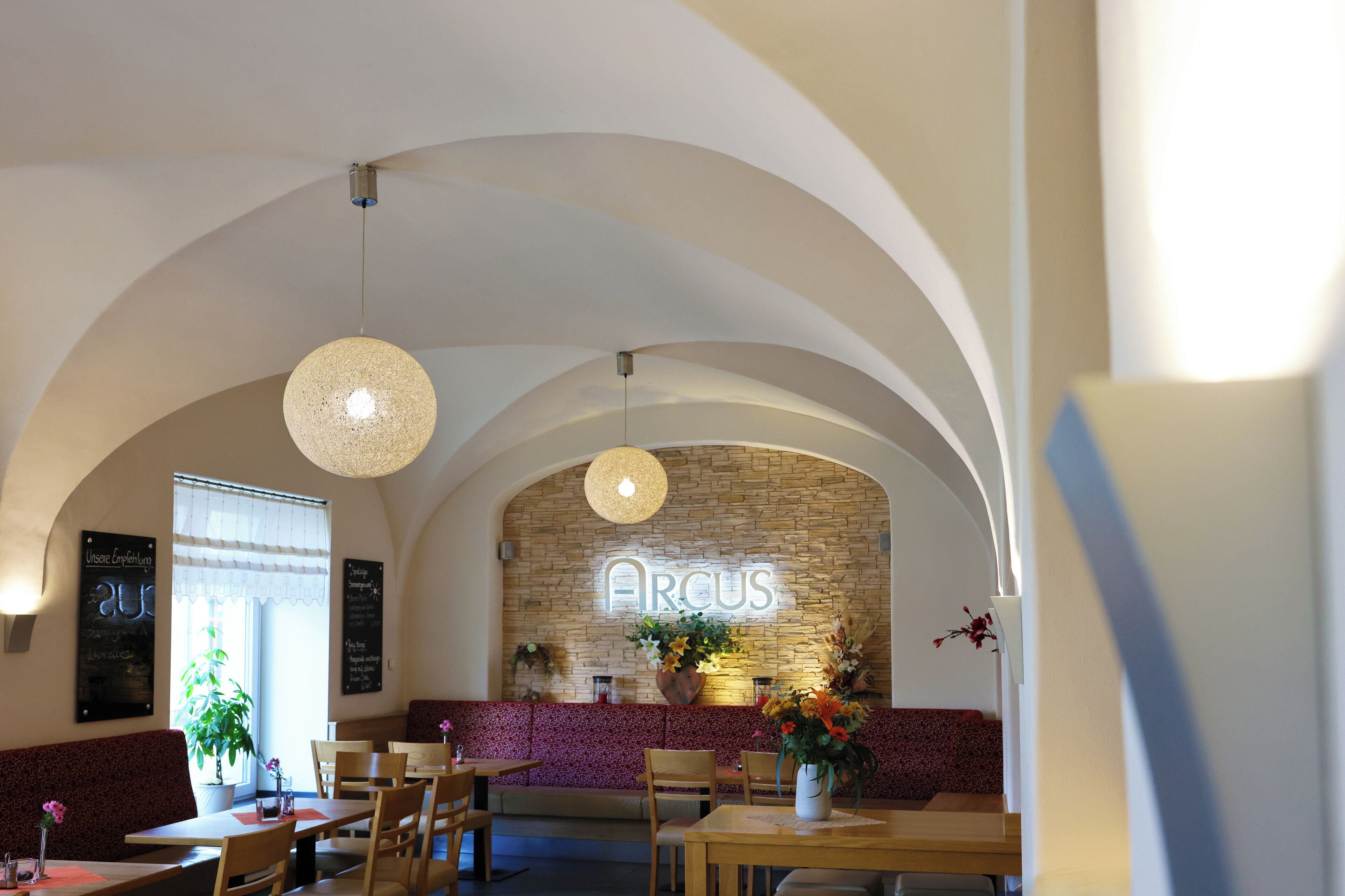 Interior de restaurante com tetos abobadados, mesas e cadeiras de madeira, um letreiro de neon com a inscrição "ARCUS" e uma grande janela com plantas.