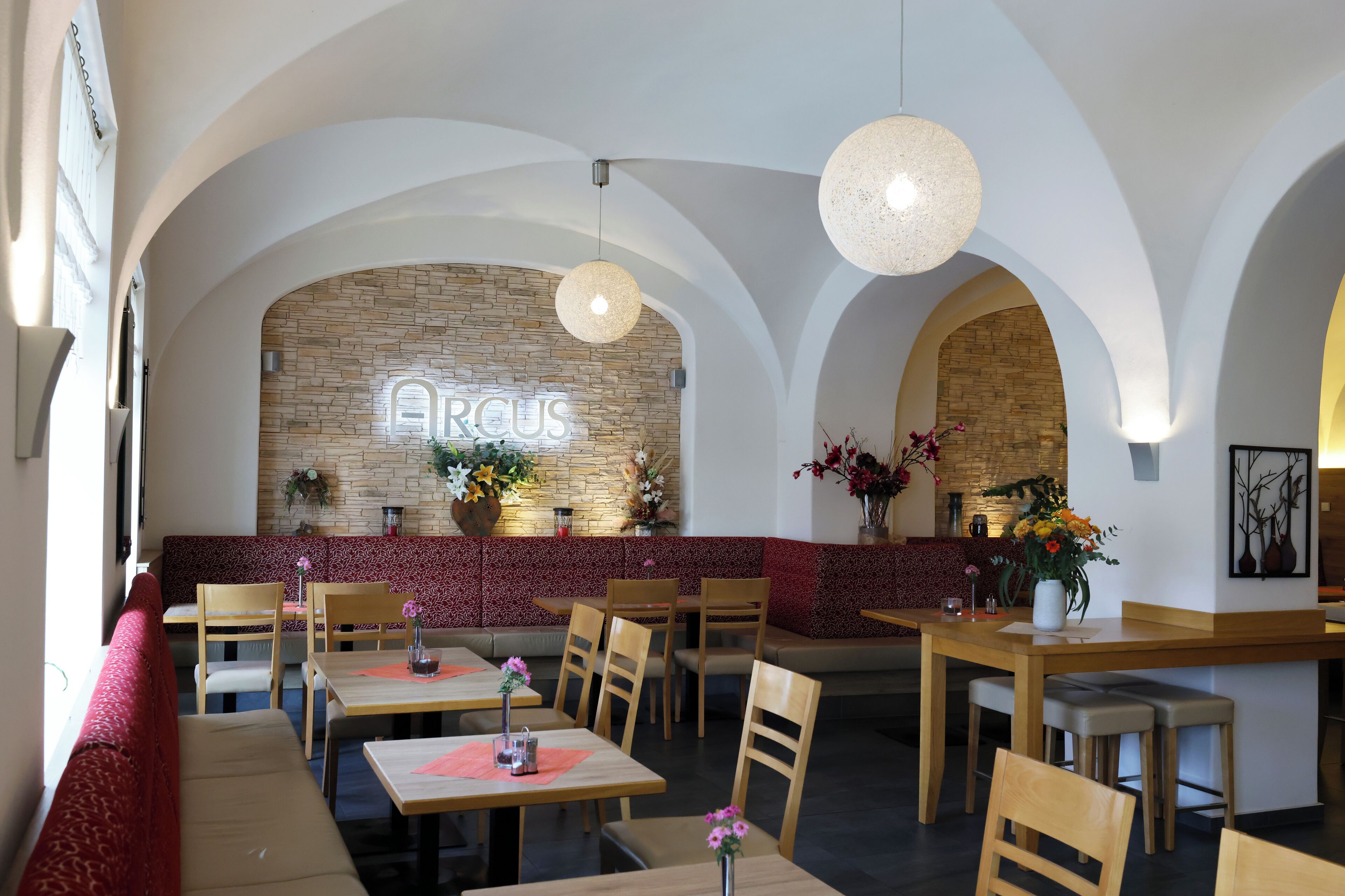 Interior de restaurante com tetos em arco, mesas e cadeiras de madeira e parede de pedra com a palavra 'ARCUS' iluminada.