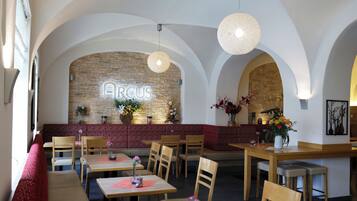 Interior de restaurante com tetos em arco, mesas e cadeiras de madeira e parede de pedra com a palavra 'ARCUS' iluminada.
