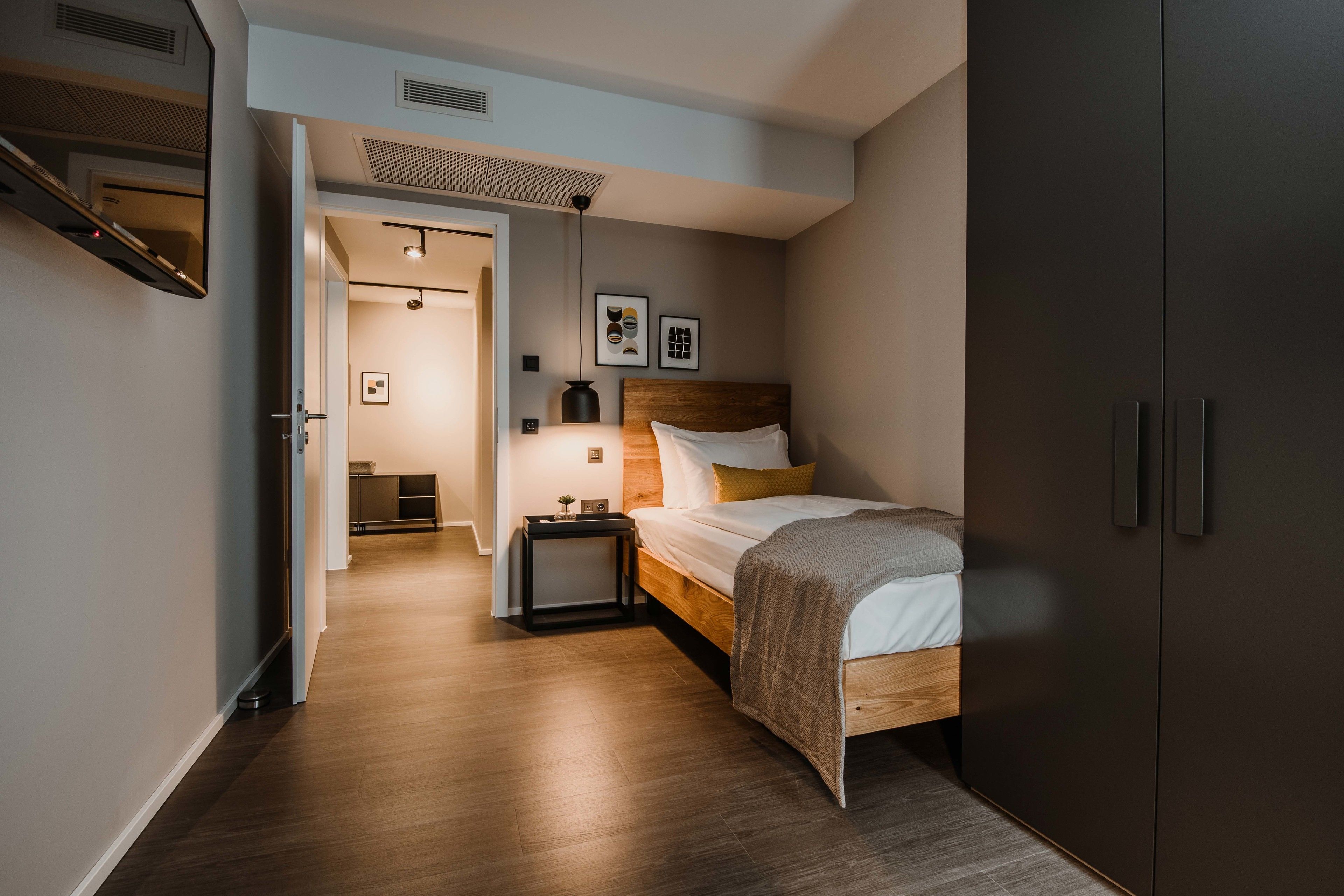 Quarto de hotel moderno com cama de madeira, mesa de cabeceira, guarda-roupa e corredor que dá para um banheiro.