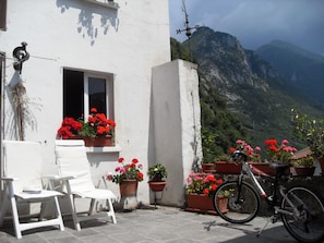 Terraço com cadeiras brancas, plantas em vasos com flores vermelhas e uma bicicleta.