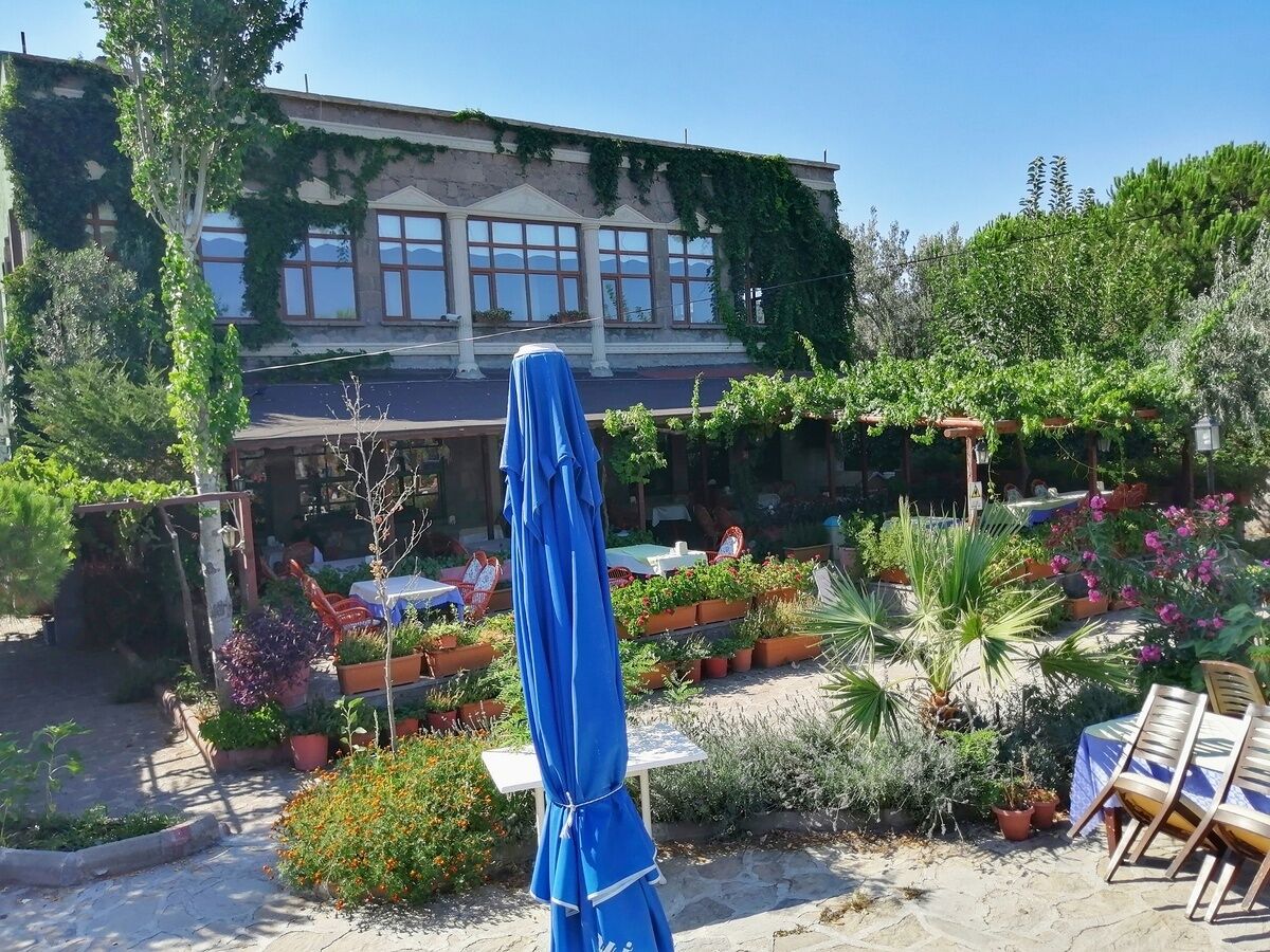 Uma área de varanda externa com mesas e cadeiras, rodeada por plantas em vasos e um edifício com janelas amplas.
