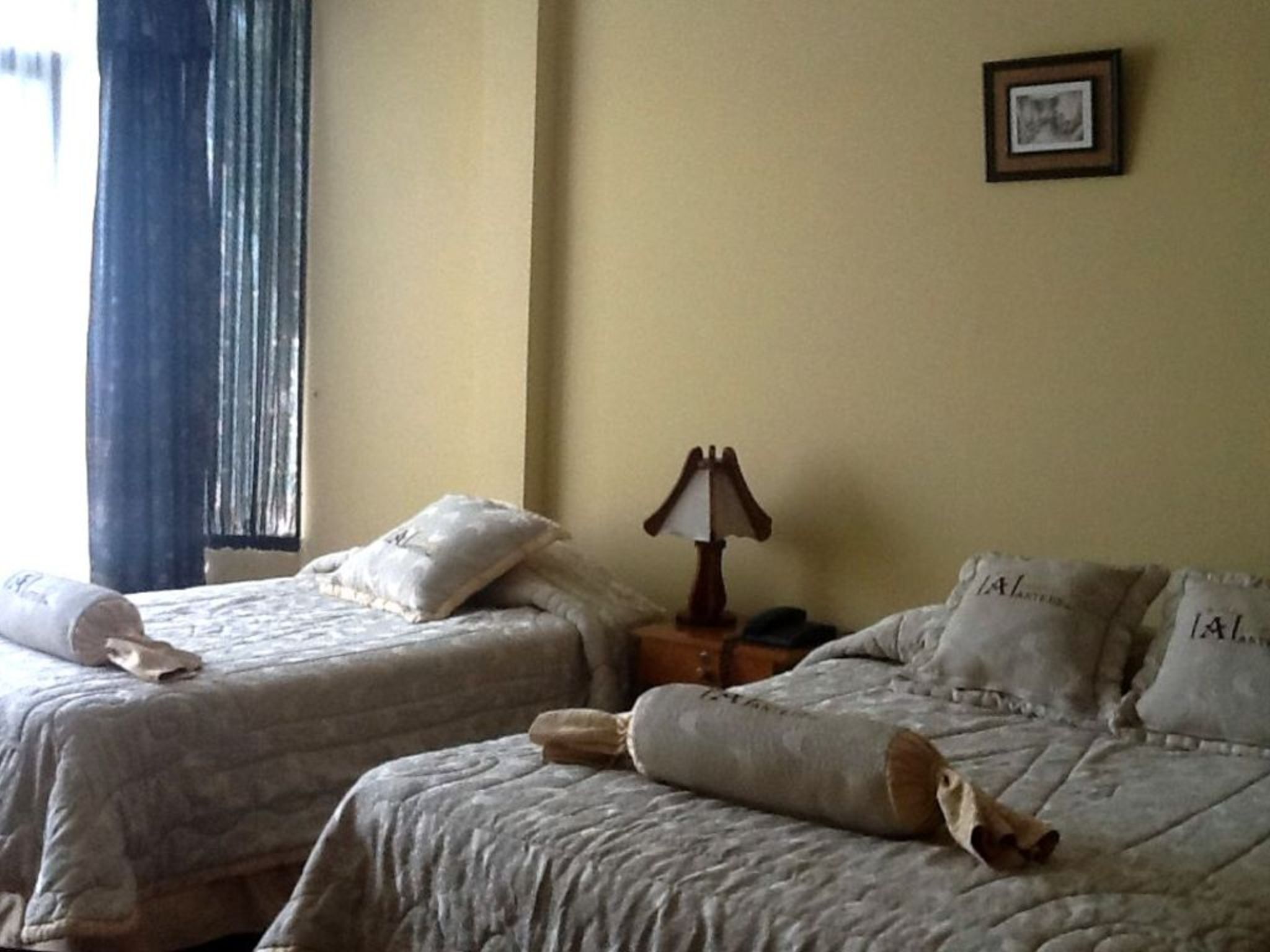 Quarto de hotel com duas camas, uma mesa de cabeceira com luminária, um quadro na parede e uma janela com cortinas.