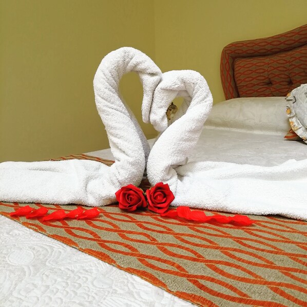 Uma cama com toalhas brancas dispostas em formato de coração e duas rosas vermelhas sobre um edredom estampado.