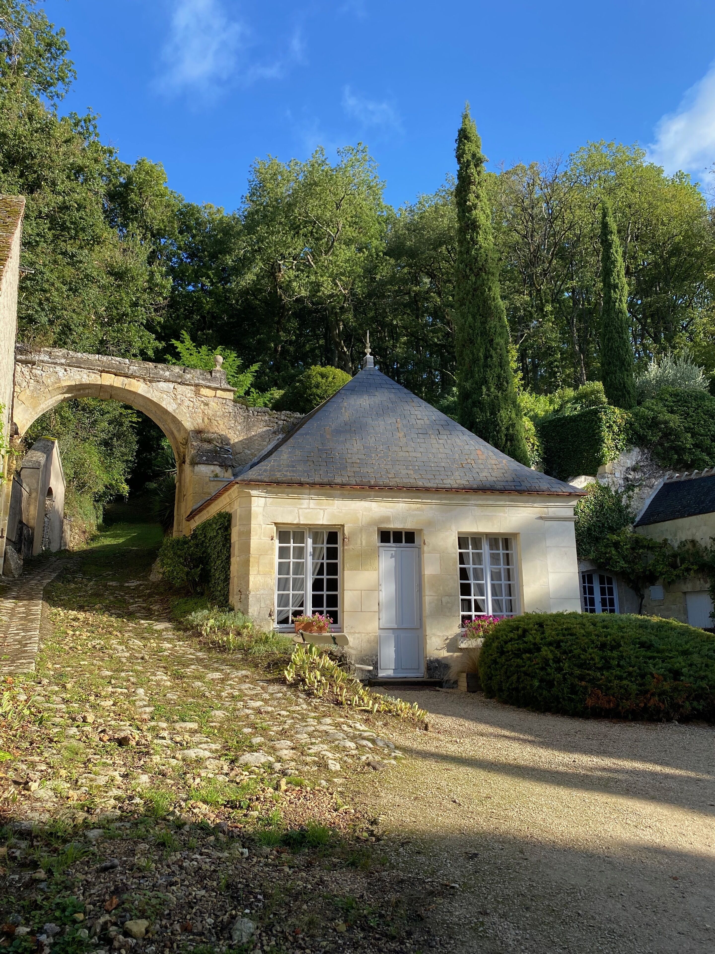 Um edifício de pedra com telhado em telhas, um arco de pedra e um caminho de cascalho que o leva até ele.