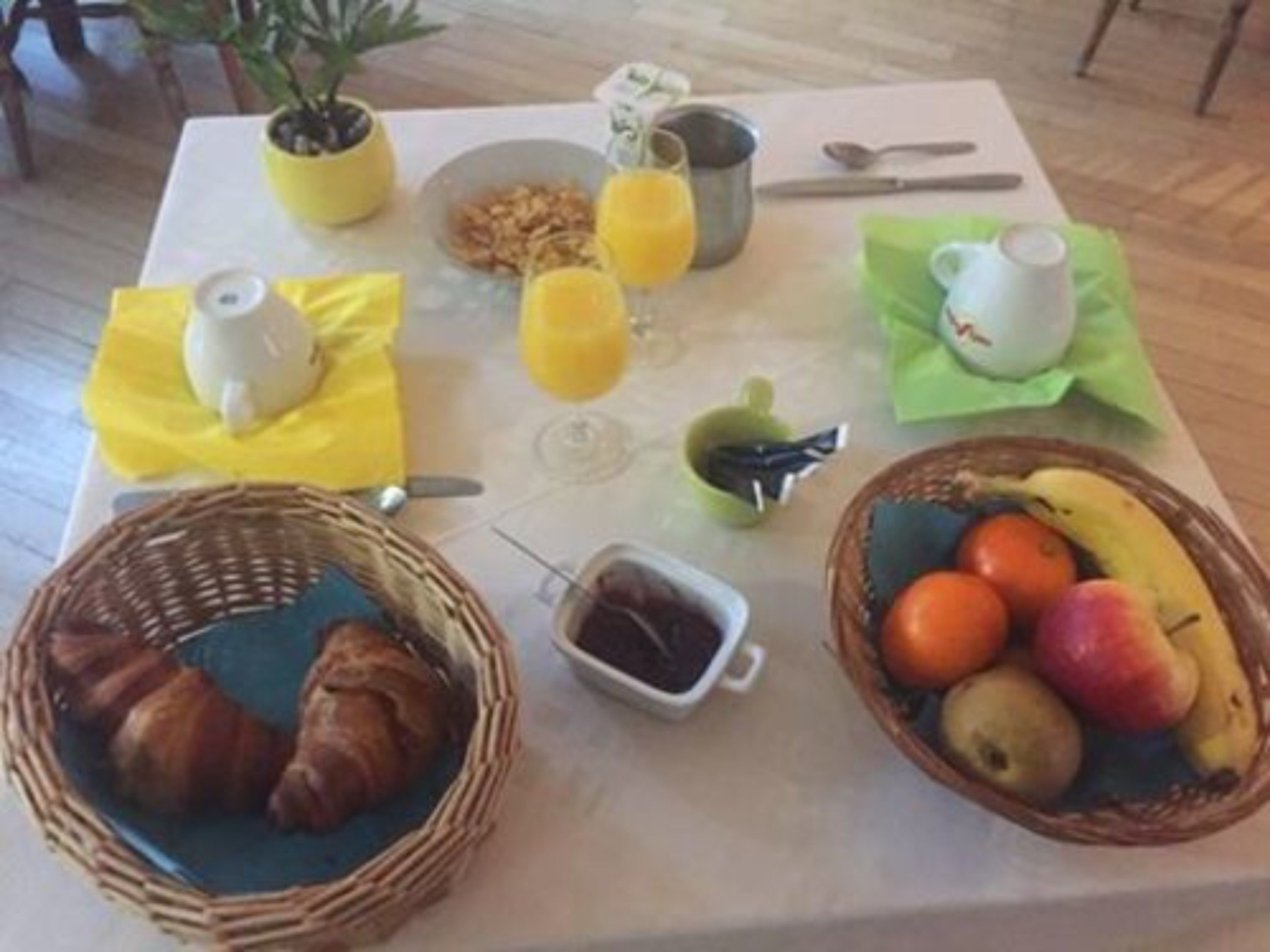 Mesa de café da manhã com croissants, uma saladeira de frutas, copos de suco e uma cafeteira.