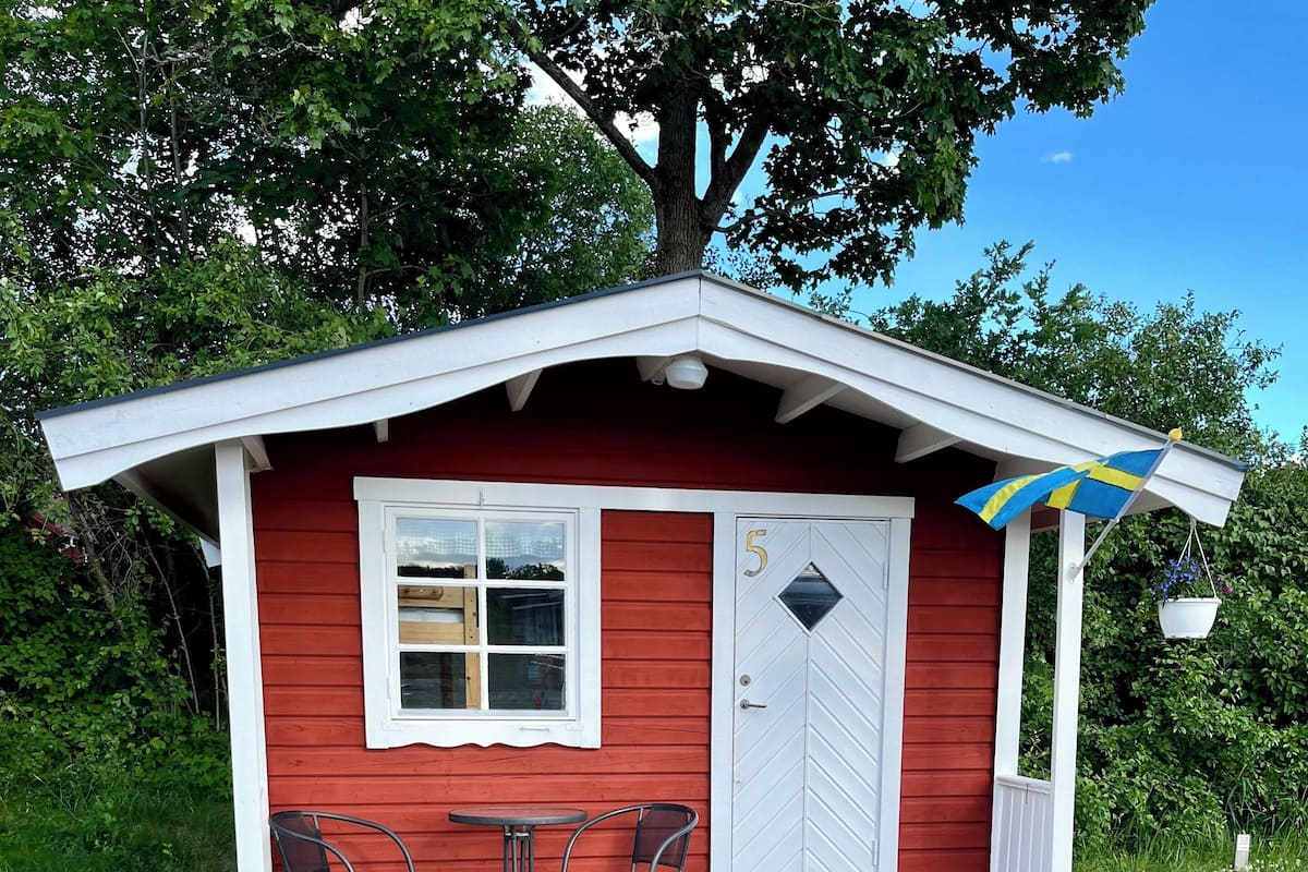 Uma pequena cabana vermelha com porta branca identificada com o número '5', uma janela e um alpendre com duas cadeiras e uma mesa.