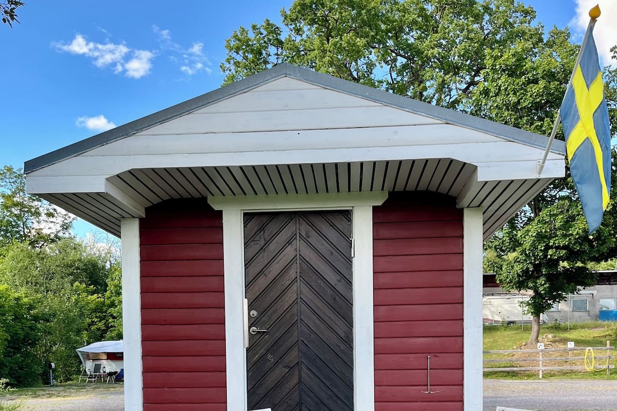 Uma pequena estrutura de madeira com exterior vermelho e porta preta, apresentando duas cadeiras dobráveis brancas e uma pequena mesa em um deck de madeira.