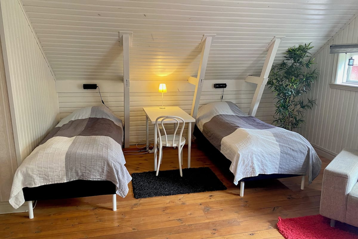 Quarto com duas camas, uma escrivaninha, uma cadeira, um abajur e uma planta.