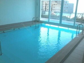 Piscina interior com água azul cristalina, escadas de metal e vista para edifícios através de grandes janelas.
