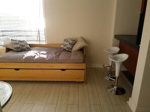 Quarto com cama de madeira, duas almofadas, uma mesa redonda pequena e um banquinho branco.