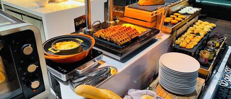 Disposição de buffet com diversos alimentos, incluindo waffles, doces e pães, dispostos em um balcão.