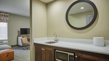 Quarto de hotel com cozinha compacta, espelho grande, TV de tela plana, sofá e janela com persianas.