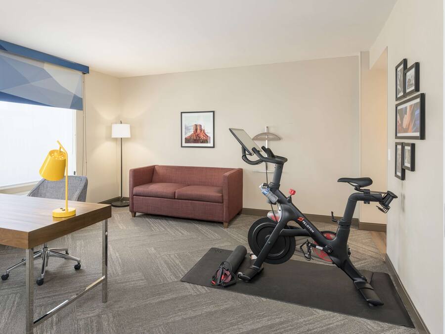 Quarto de hotel com secretária de madeira, sofá, luminária de mesa, bicicleta, bicicleta ergométrica fixada na parede e uma moldura com uma imagem.