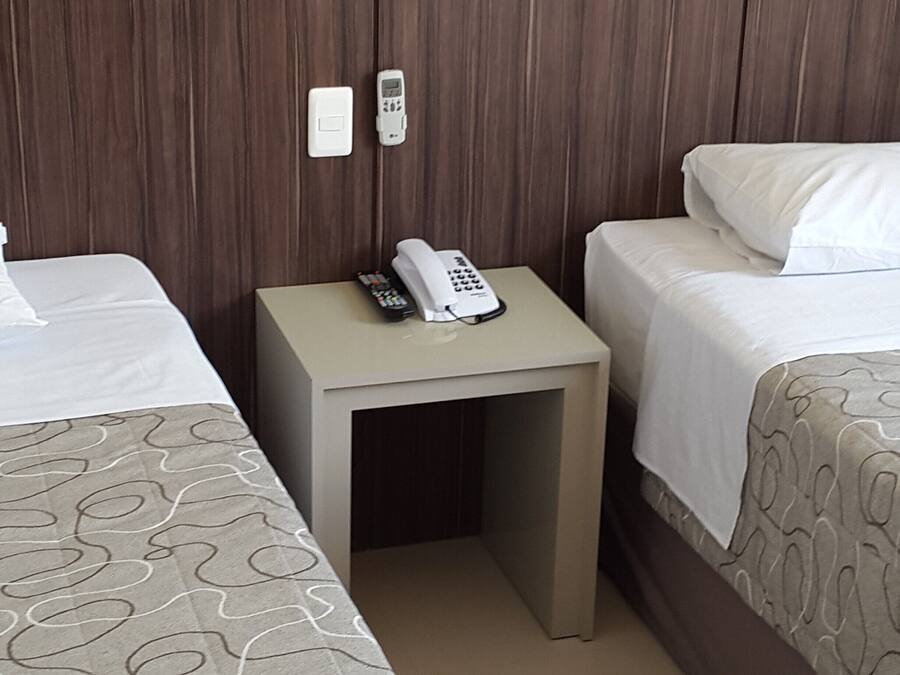 Quarto de hotel com duas camas, cada uma com travesseiro branco e colcha estampada. Entre as camas, há uma mesinha com um telefone e um controle remoto.