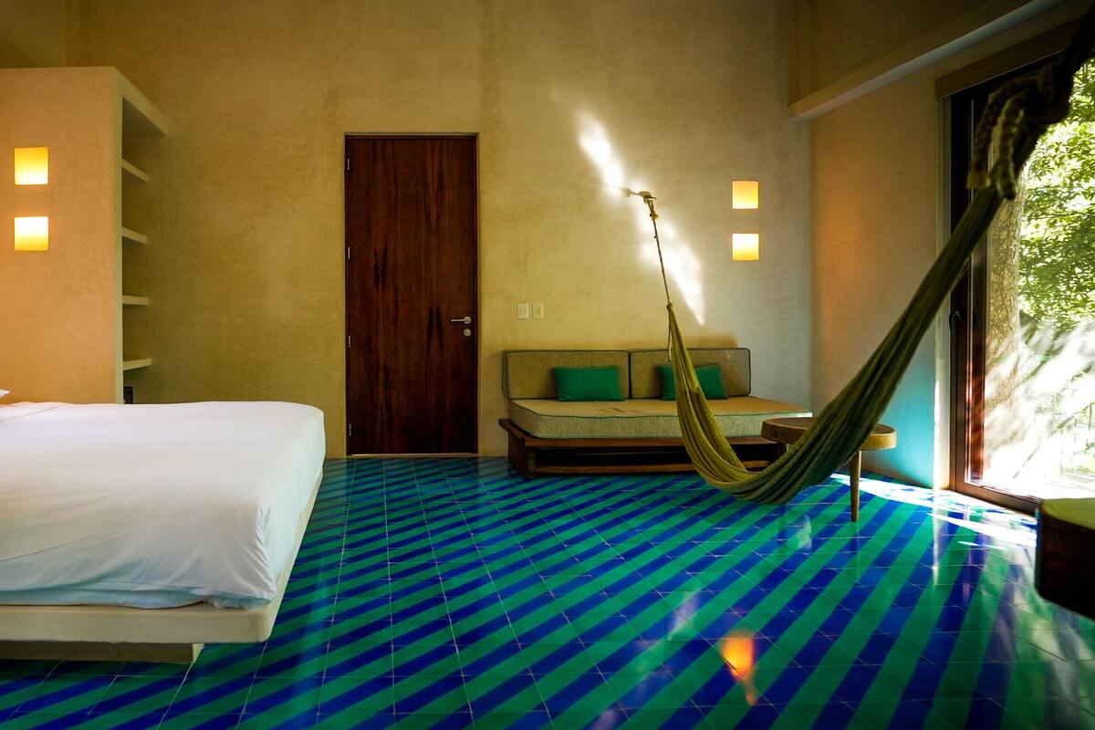 Quarto de hotel com cama, sofá, rede e uma janela grande.