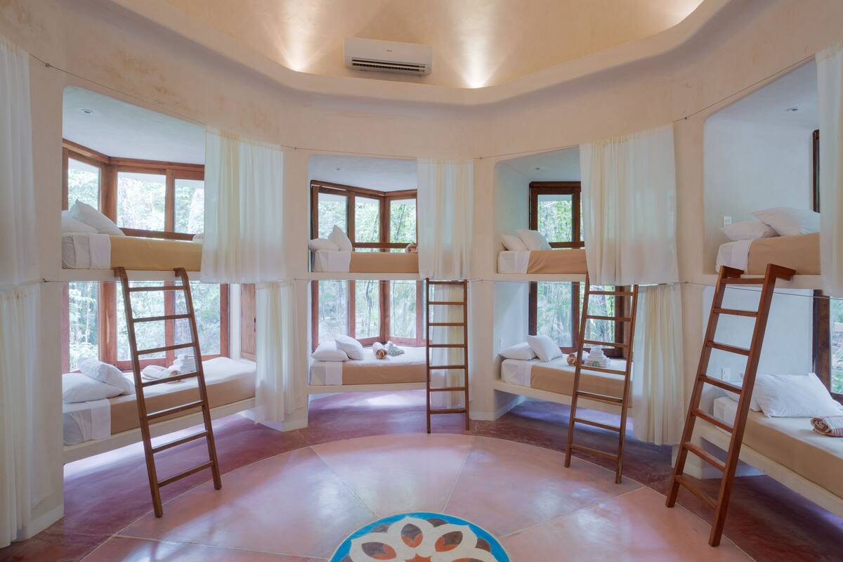 Quarto circular com beliches, escadas de madeira e janelas amplas.