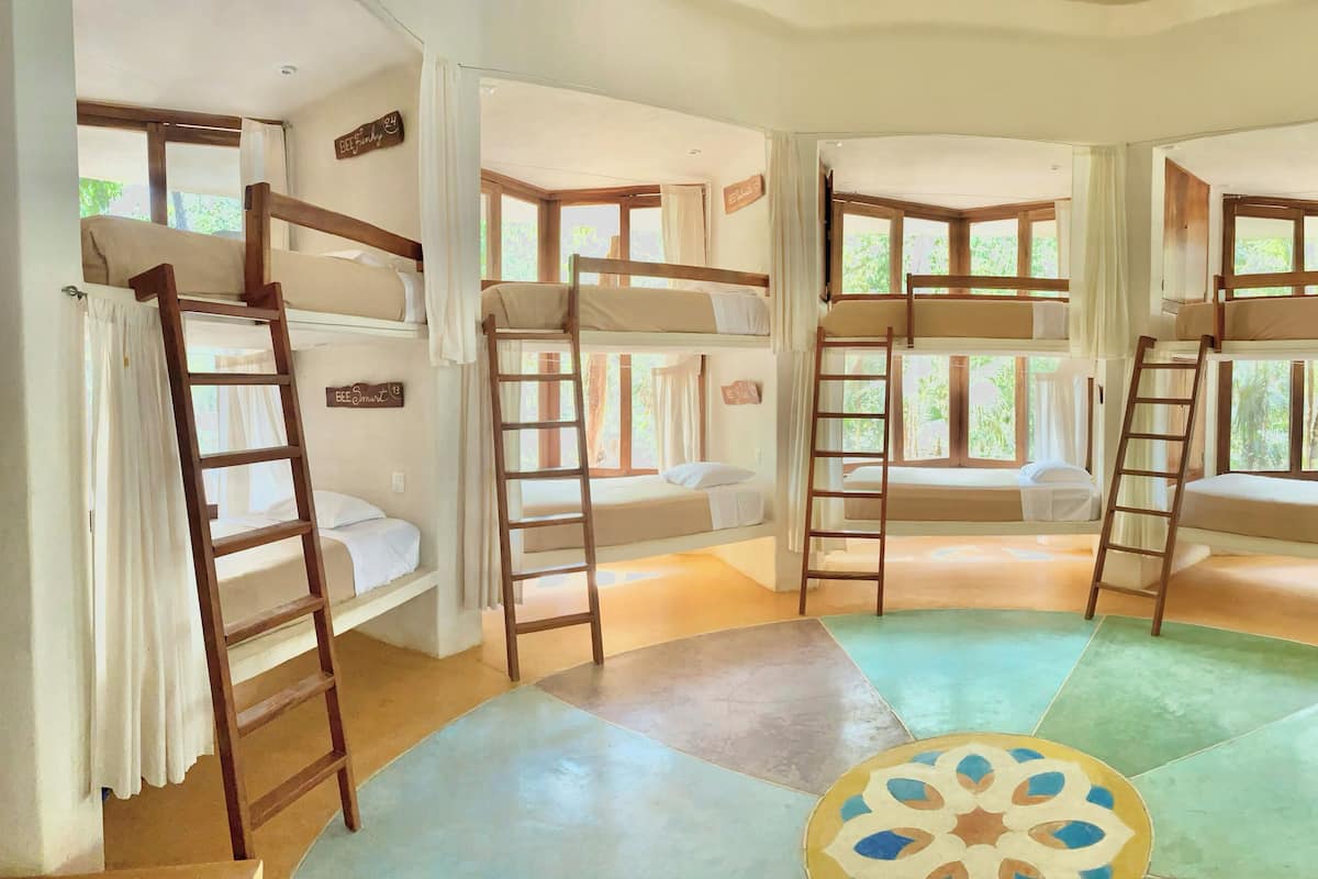 Quarto circular com beliches e escadas, paredes de madeira e janelas grandes.