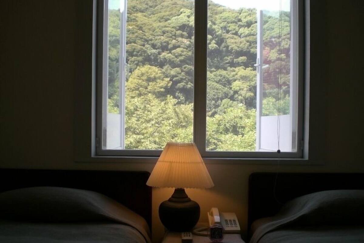 Quarto de hotel com cama, luminária, telefone e janela com vista para uma floresta.