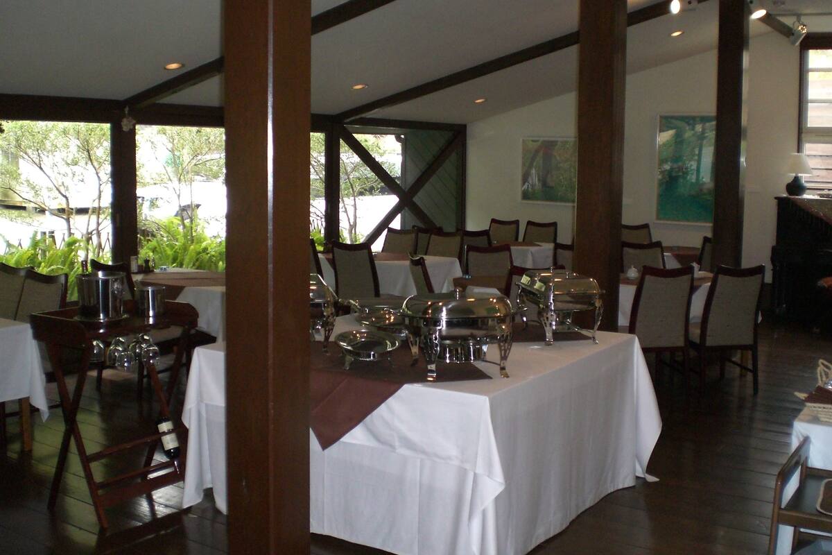 Uma sala de jantar com mesas preparadas para uma refeição, vigas de madeira no teto e vista para o verde lá fora.