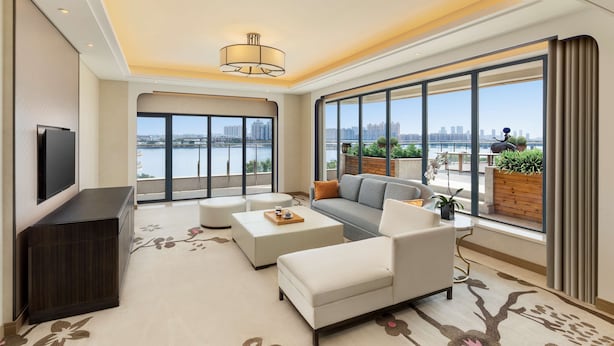 Uma sala de estar moderna com um sofá, duas poltronas brancas, uma mesa de centro e uma TV sobre um móvel escuro. Janelas amplas oferecem vista para o horizonte urbano e um corpo d'água.
