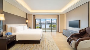 Quarto de hotel moderno com uma cama grande, uma cadeira de massagem, uma TV de tela plana e vista para a cidade através de janelas amplas.