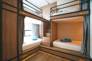 Quarto com beliches, paredes de madeira e janela com cortinas. Há duas camas, uma no beliche superior e outra no beliche inferior.