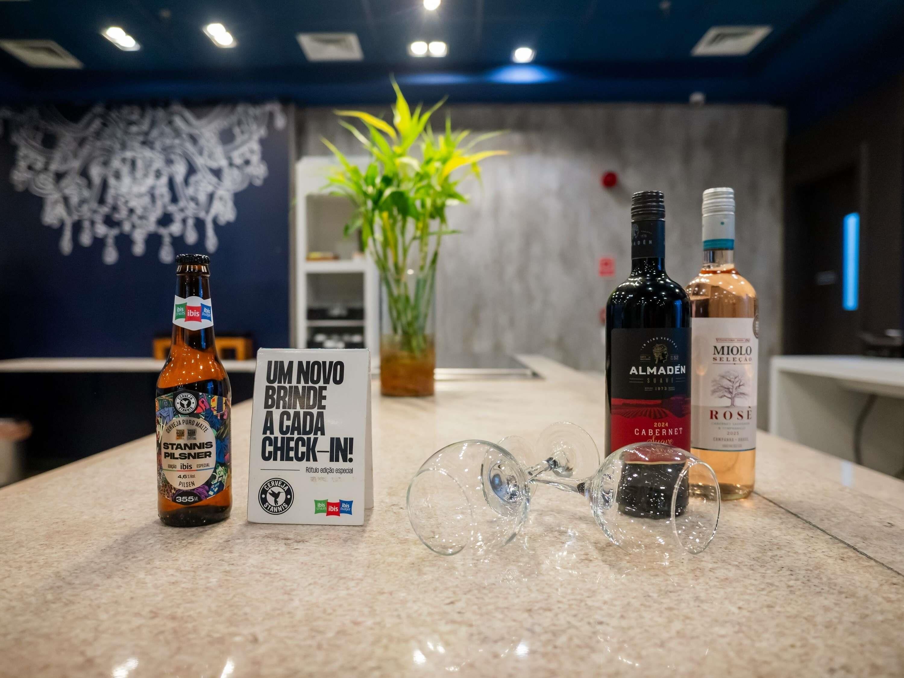 Uma área de bar com uma garrafa de cerveja Stannis, uma garrafa de vinho Almodéen, uma garrafa de vinho rosé Mielo Seleção, e um letreiro com a frase 'UM NOVO BRINDE A CADA CHECK-IN'.