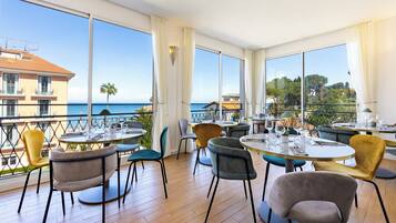 Uma sala de jantar com mesas e cadeiras redondas, janelas amplas com cortinas e vista para o mar e edifícios.