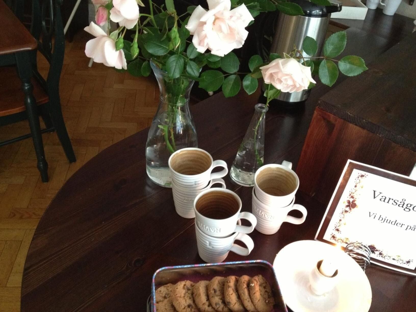 Uma mesa com chávenas de café, um prato de bolachas e um vaso com rosas.