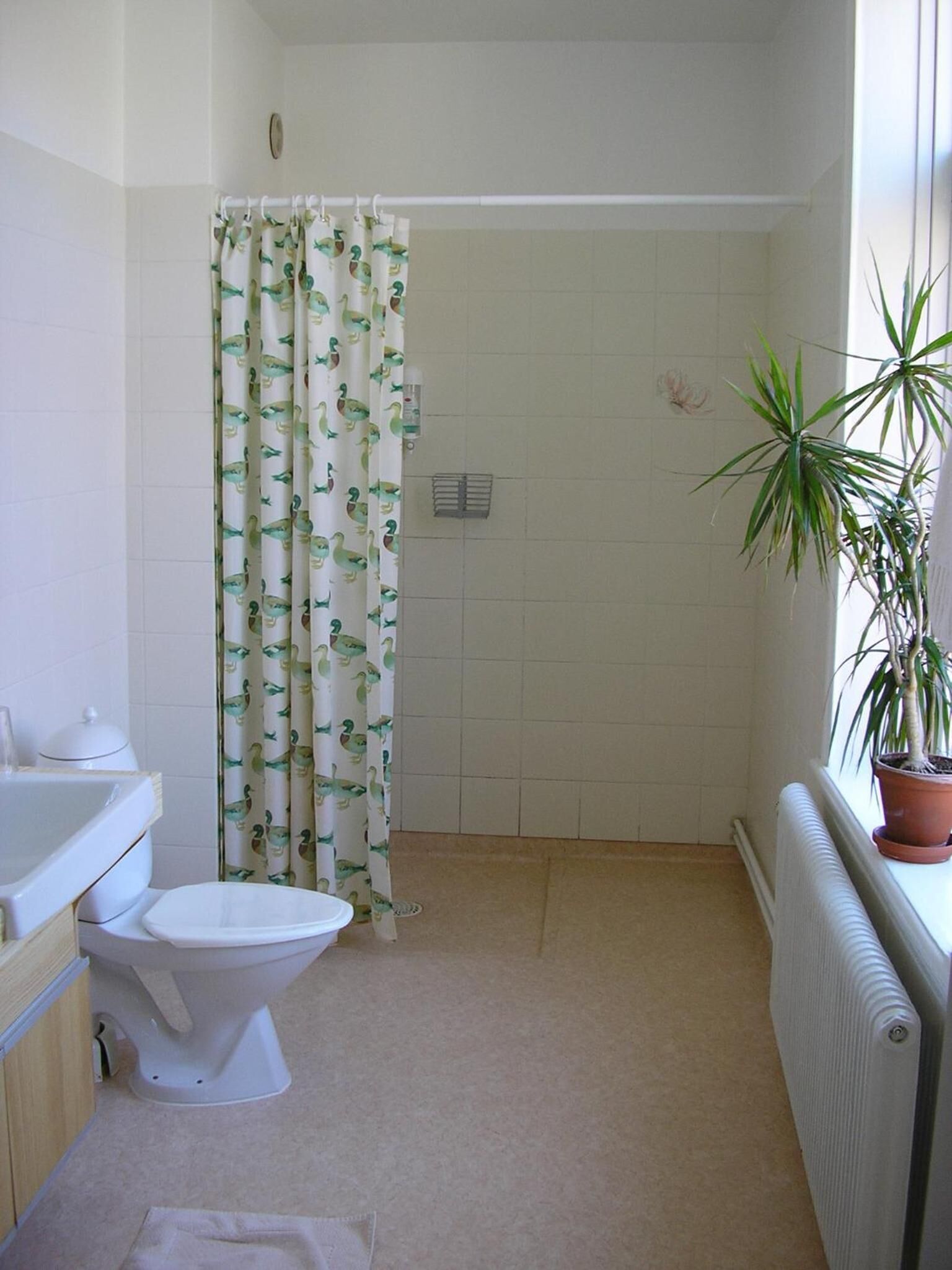Uma casa de banho pequena com sanita, cortina de chuveiro com padrão verde e branco, e uma planta num vaso perto da janela.