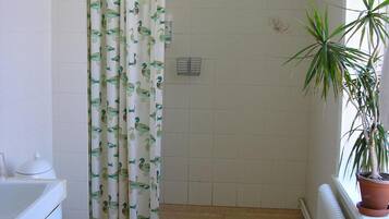 Uma casa de banho pequena com sanita, cortina de chuveiro com padrão verde e branco, e uma planta num vaso perto da janela.