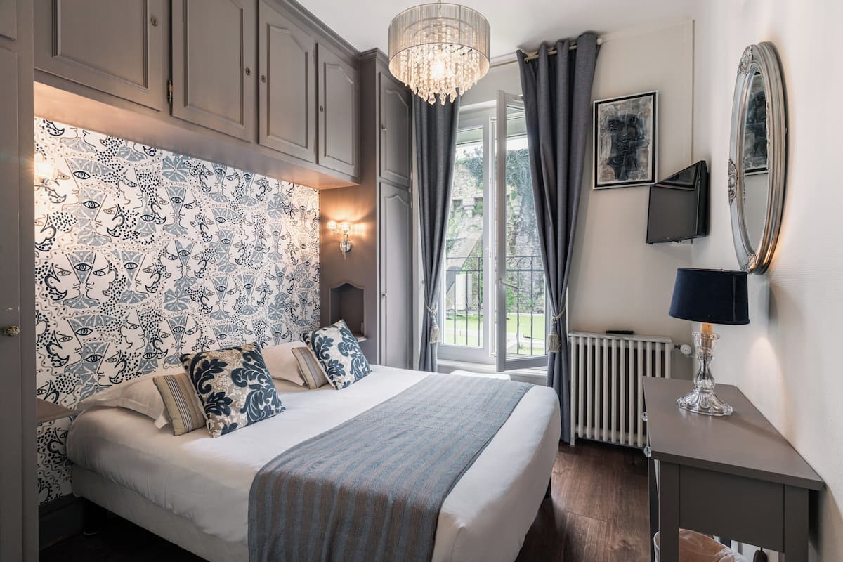 Quarto com cabeceira estampada, cama com vários travesseiros, mesa de cabeceira com luminária, espelho e janela com cortinas.