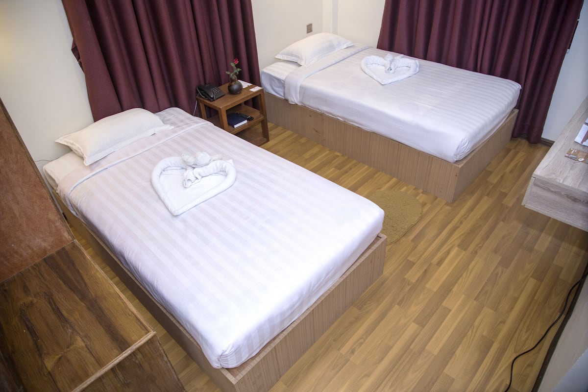 Quarto de hotel com duas camas, piso de madeira e uma pequena mesa de cabeceira com abajur e um livro.