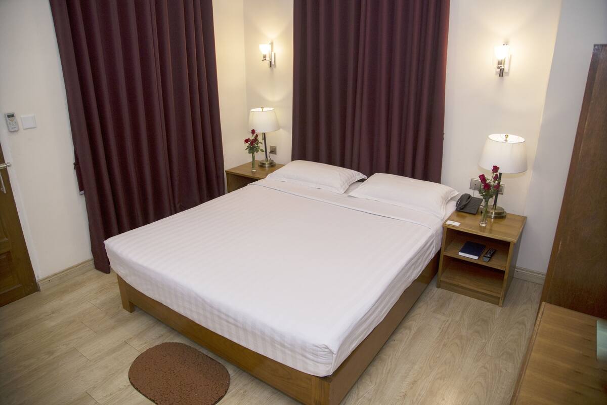 Cama bem arrumada com lençóis brancos, duas mesas de cabeceira com luminárias e um vaso de flores, e piso de madeira.