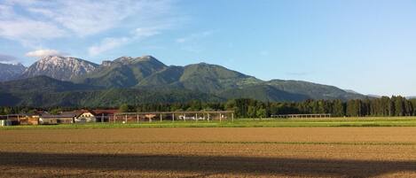 Paisagem rural com um campo, um edifício de telhado vermelho e uma cadeia de montanhas ao fundo.