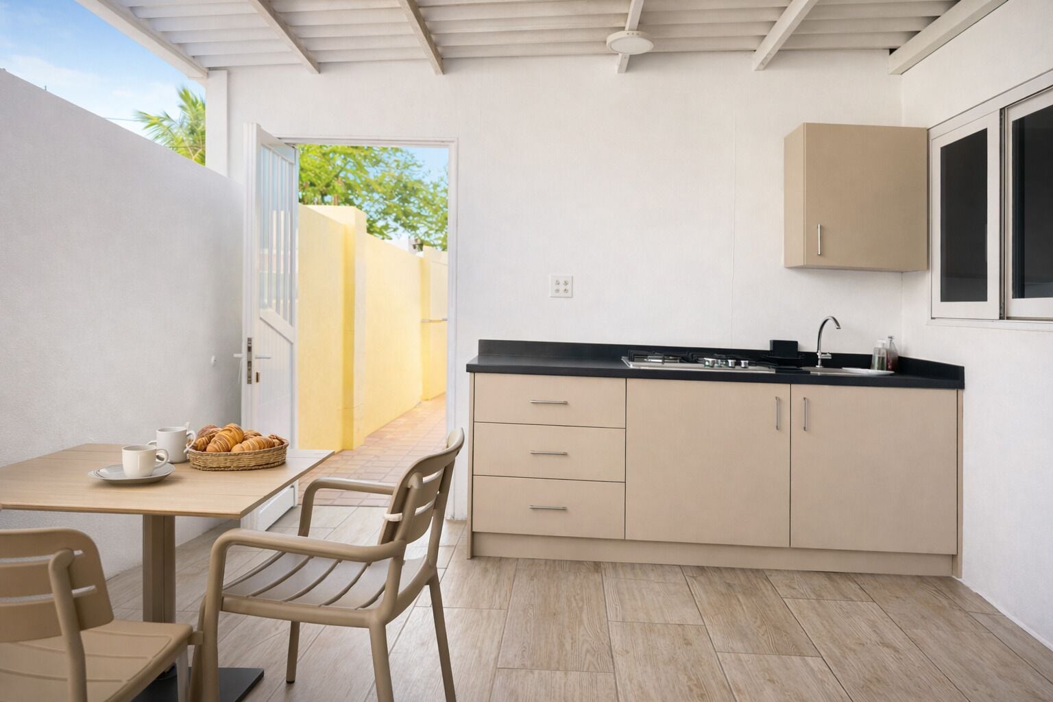Cozinha compacta com área de refeições, com mesa e cadeiras, cesta de doces e vista de um prédio amarelo através de uma porta aberta.