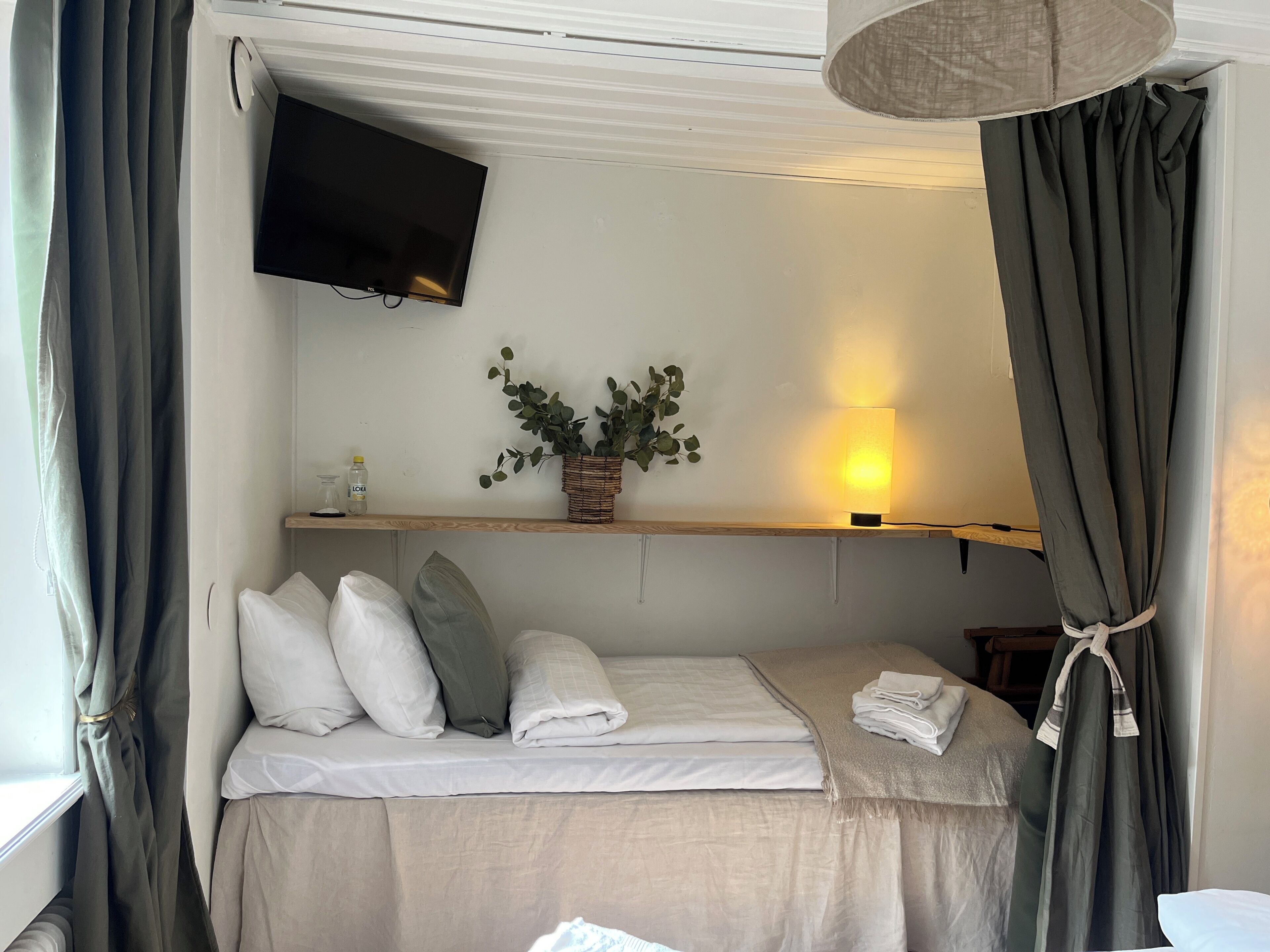 Quarto de hotel com cama, televisão na parede, prateleira com uma planta e abajur.