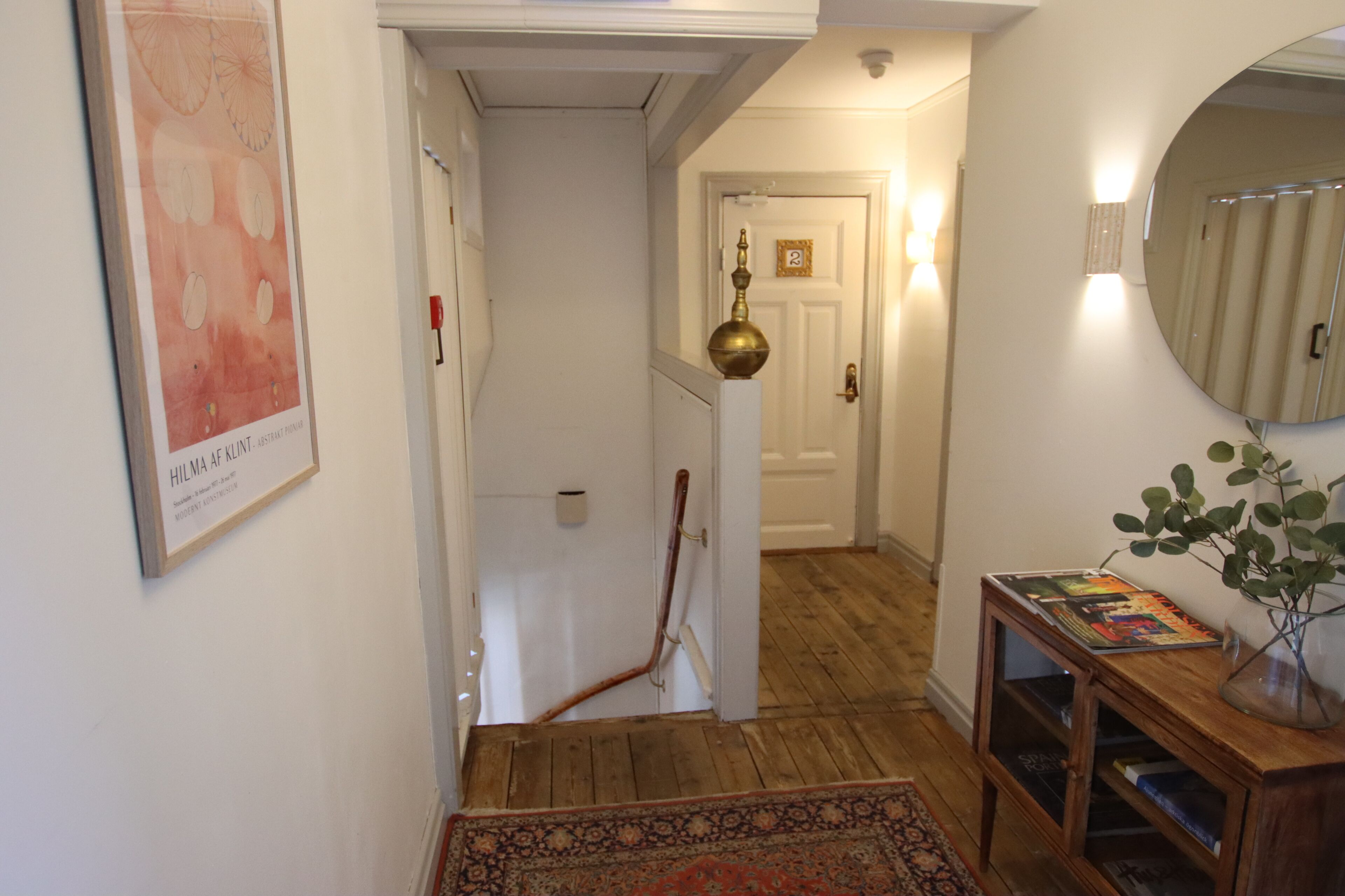 Um corredor com piso de madeira, tapete estampado, mesa consola de madeira com um vaso e um quadro na parede.