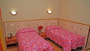 Um quarto pequeno e acolhedor com duas camas, uma mesinha de cabeceira, um vaso com flores e duas luminárias na parede.