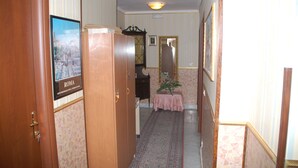 Um corredor com uma porta de madeira, um cartaz de Roma, um piano, uma pequena mesa com toalha rosa e um espelho na parede.