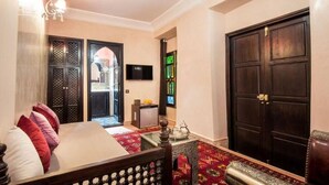 Quarto com cama, almofadas, televisão, mesinha, espelho decorativo e uma grande porta de madeira.