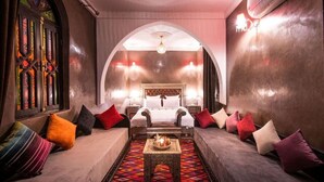 Quarto de hotel luxuoso com uma cama grande, duas mesas de cabeceira, tapete estampado e almofadas coloridas.