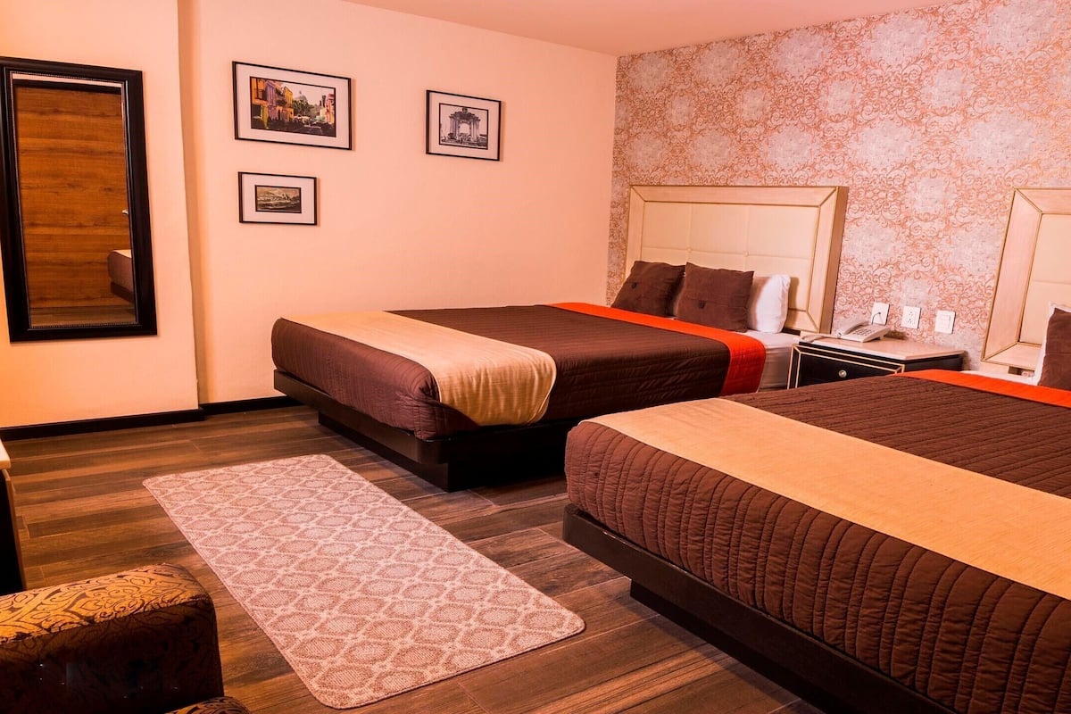 Quarto de hotel com duas camas, um pequeno sofá, um tapete estampado e uma decoração na parede.
