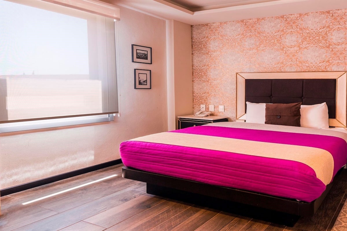Quarto de hotel com cama, mesa de cabeceira, janela e parede estampada.