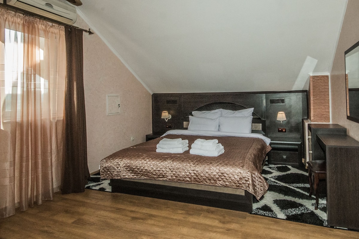 Quarto de hotel com uma cama grande, teto de madeira, mesa de cabeceira com abajur e escrivaninha com cadeira.